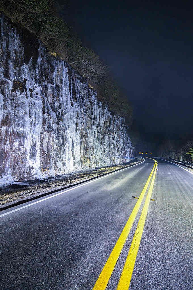 Winter ice formations cling to rock faces, creating natural sculptures illuminated by headlights on evening drives.