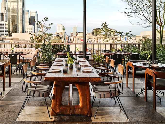 A communal wooden table invites strangers to become friends over shared plates and city panoramas.