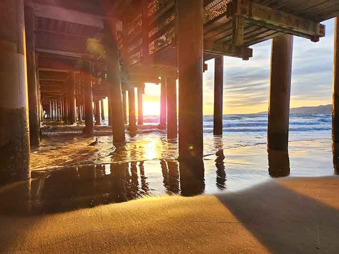 Golden sunset light streams through weathered pier pilings while waves dance beneath the vintage amusement park's cheerful glow.