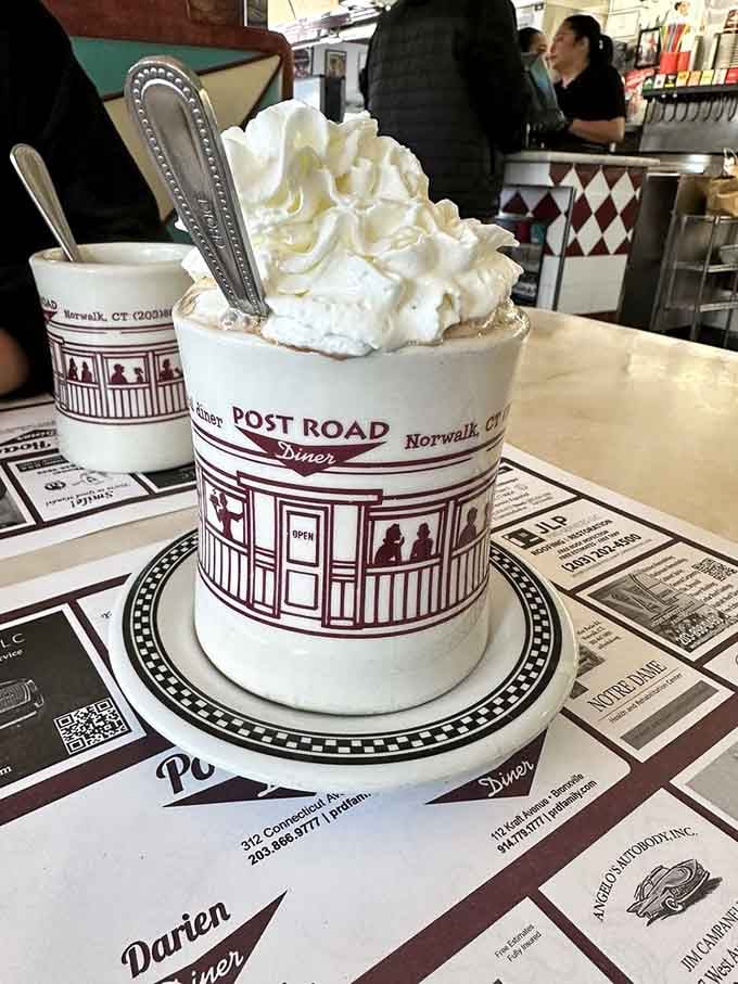 Hot chocolate piled high with whipped cream in a branded mug, because sometimes you need dessert disguised as beverage.