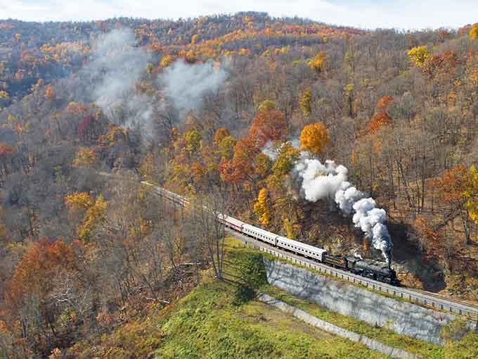 An aerial view captures the steam train winding through fall foliage, looking like a postcard that somehow came to life.
