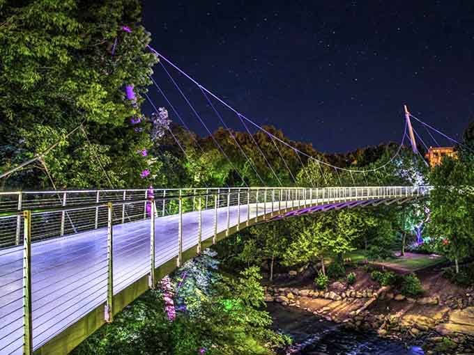 The Liberty Bridge glows like a purple ribbon at night, suspended gracefully above the rushing Reedy River below.
