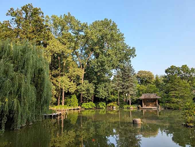 Graceful willows drape over tranquil waters where a traditional tea house sits peacefully among towering trees and manicured greenery.