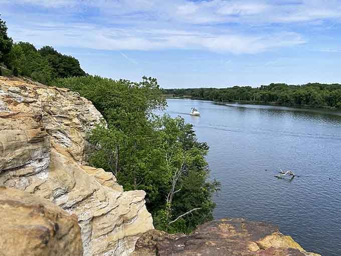 Summer greenery hugs the cliff edge where boats drift lazily below, completely unaware of the drama above.