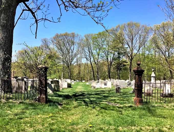 Ornate iron gates frame this peaceful spring scene where weathered headstones stand among fresh grass and budding trees.