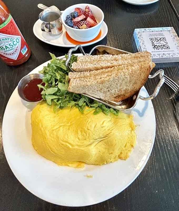 That omelet stands tall and proud like a golden pillow of perfection, flanked by toast and fresh fruit companions.
