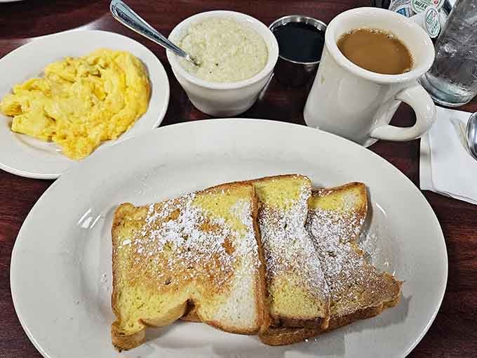 French toast dusted with powdered sugar sits alongside scrambled eggs and grits, the breakfast trinity that never disappoints anyone.