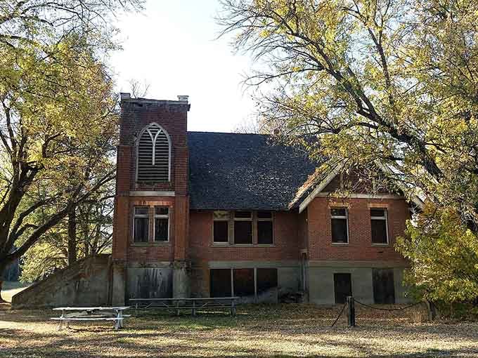 The weathered brick church stands silent among golden autumn leaves, its empty windows watching over forgotten memories.