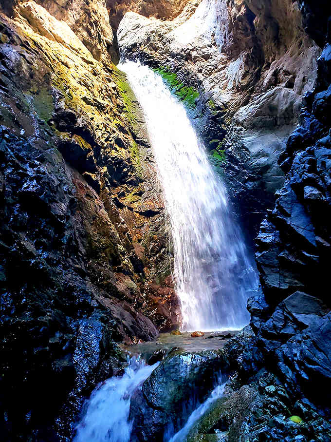 Sunlight catches the rushing water as it plunges through this narrow slot canyon into a misty pool below.