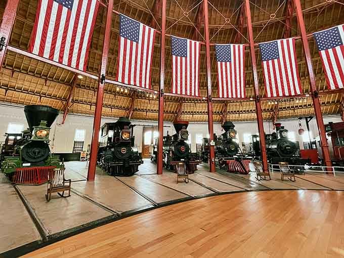 Five magnificent locomotives stand beneath soaring flags in this cathedral-like roundhouse, a testament to American railroad heritage.