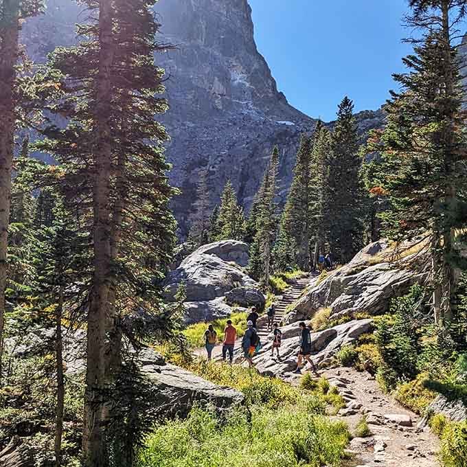 Families climb the rocky trail together, dwarfed by the massive cliff face that looms dramatically overhead.