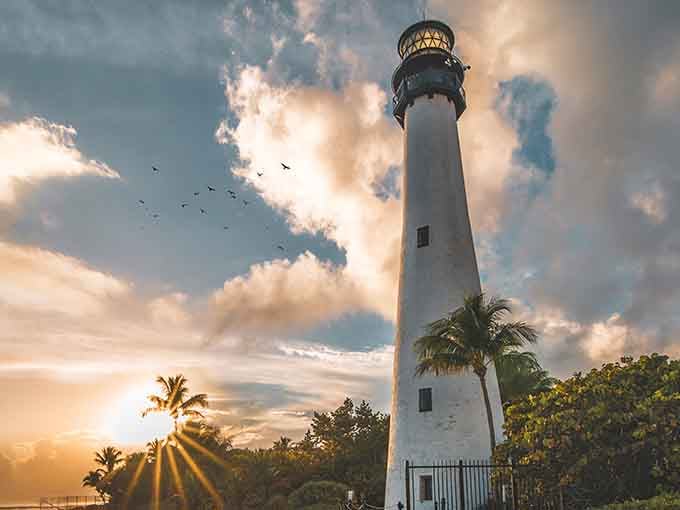 Dramatic sunset light breaks through clouds, painting the sky while birds circle this majestic coastal guardian in golden hour glory.