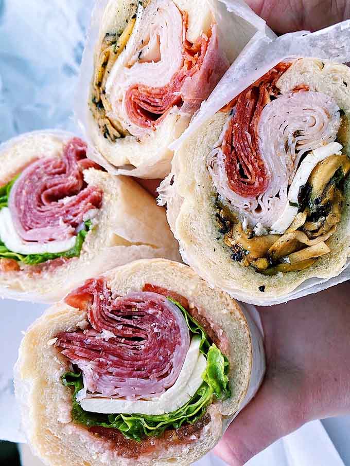 Cross-sections revealing layers of Italian cold cuts stacked like delicious architecture. This is sandwich engineering at its finest level.