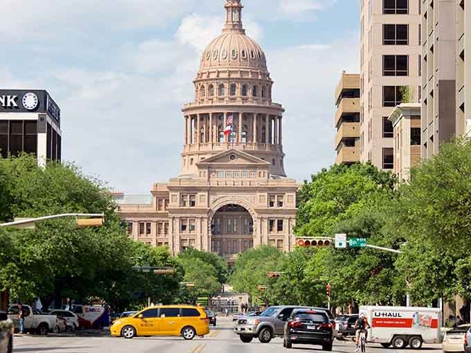 That pink granite capitol dome rises majestically through the trees, a stunning reminder of Texas pride framed by leafy green branches and blue sky.
