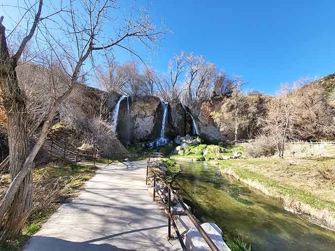 Twin waterfalls cascade down the limestone cliff while bare winter trees frame this peaceful paved pathway below.