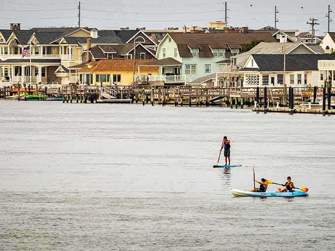 Colorful beach houses reflected in calm waters while paddleboarders glide by—summer vacation goals achieved perfectly here.
