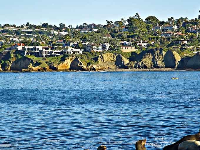 Dramatic cliffs cradle elegant homes perched above sapphire waters where kayakers glide past like tiny water bugs.