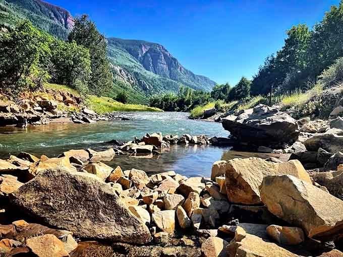 Crystal-clear river water tumbles over smooth stones, with mountains rising majestically beyond this natural paradise.