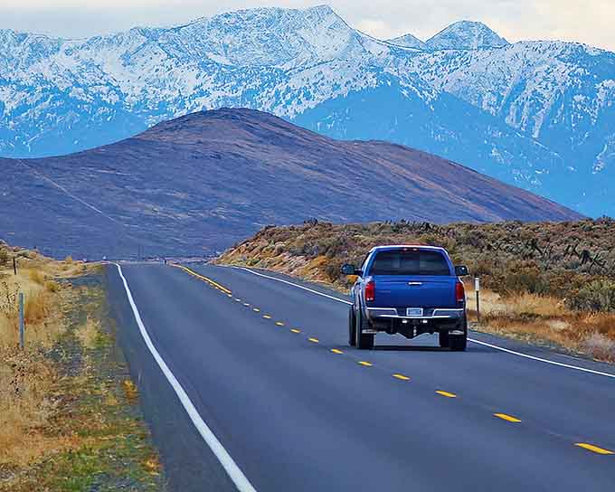 Snow-capped peaks rise beyond the highway as a lone truck heads toward mountains that touch the clouds.