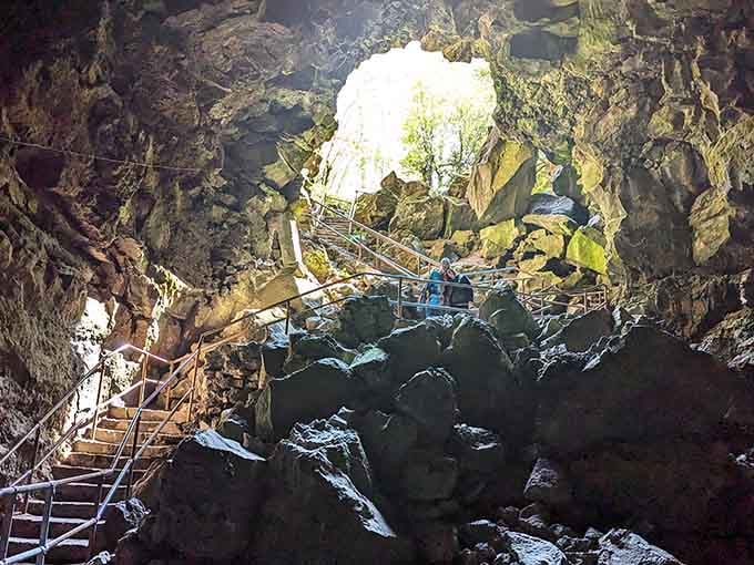 Sunlight streams through the cave opening above metal stairs, illuminating rocks in golden hues worth the climb down.