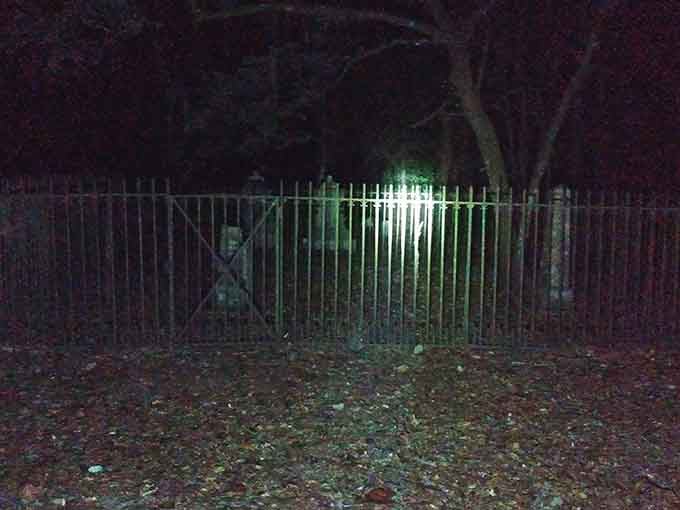 Flashlight beams pierce the darkness around iron gates, revealing shadows that daylight mercifully conceals from casual visitors.