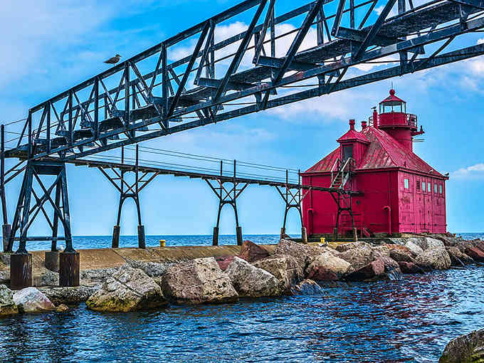 That crimson lighthouse stands bold against blue water, its catwalk stretching like an iron bridge connecting land to maritime history and adventure.