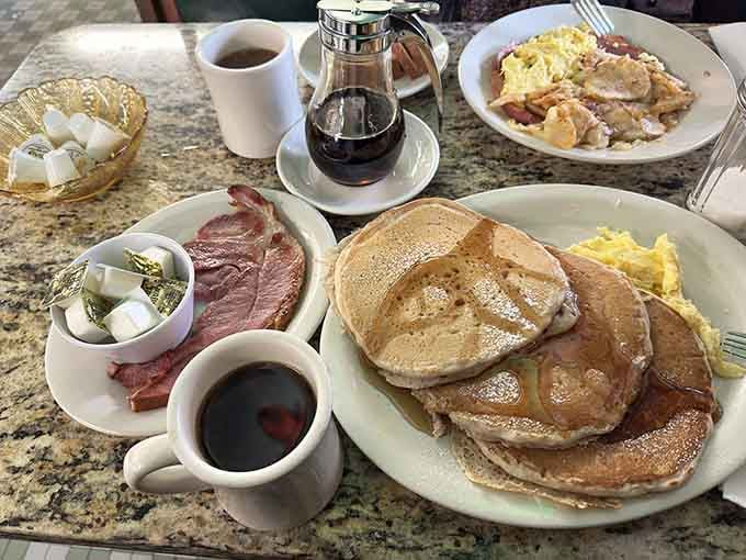 Stacks of pancakes tower beside ham and eggs while coffee flows freely, breakfast done absolutely right.