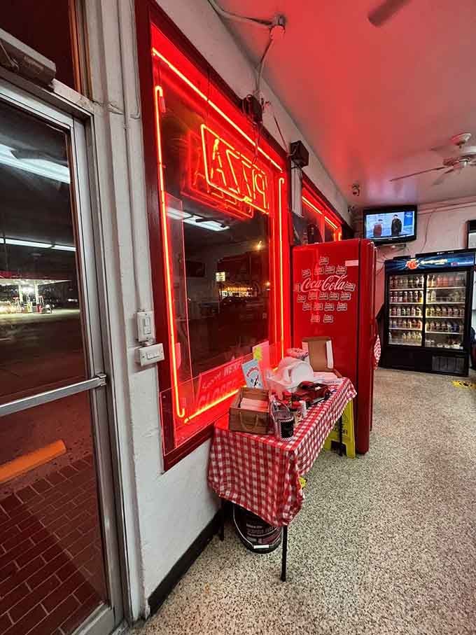 Neon "PIZZA" glowing red like a delicious warning sign, with cold drinks waiting patiently in their glass fortress.