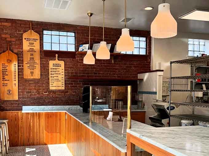 Exposed brick, warm wood, marble counters, and pendant lights create an atmosphere where bread becomes an art form.