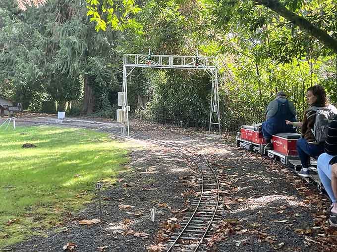 Passengers aboard a bright red locomotive cruise beneath a miniature signal gantry, living their best tiny train life.