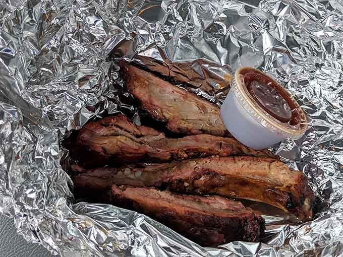 Smoke-kissed ribs resting in foil with sauce on the side, ready to make your taste buds very happy.
