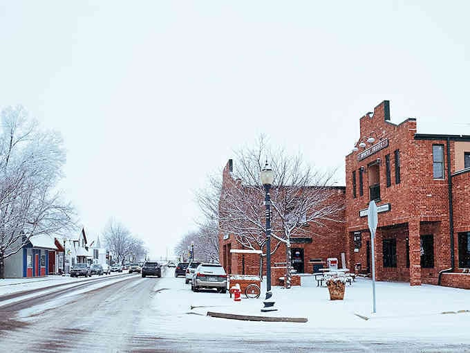 Fresh snow transforms brick buildings into a Currier and Ives print where winter's hush settles over every storefront and sidewalk.