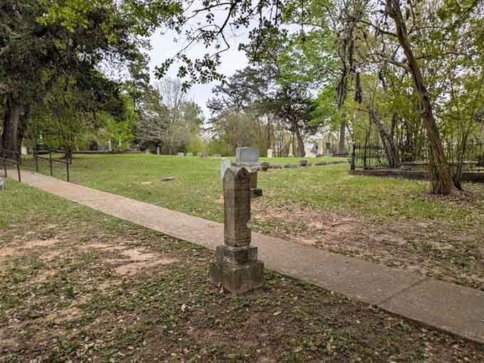 A solitary weathered marker stands beside the walking path, framed by towering trees that have witnessed decades of change.