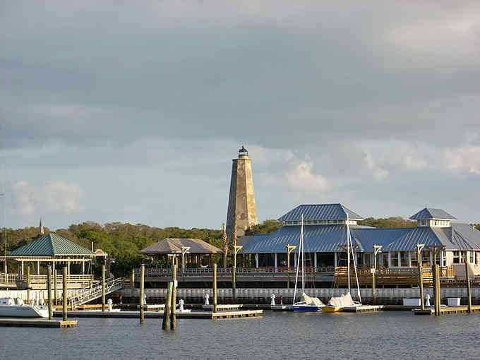 The lighthouse rises behind waterfront buildings where boats dock and seafood dinners await under coastal Carolina skies.