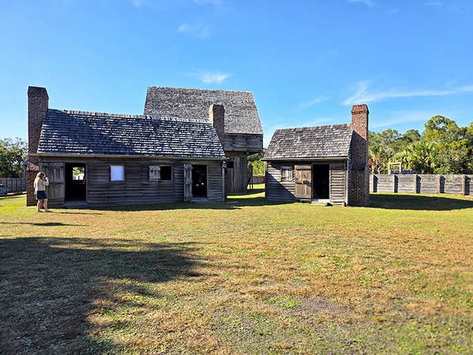 Weathered wooden buildings stand exposed under bright sun, their dark timber and pointed stakes evoking frontier danger and hardship.