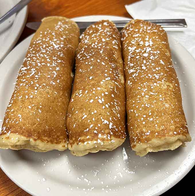 Three golden pancake rolls dusted with powdered sugar, proving that wrapping sausages in pancakes was genius innovation.