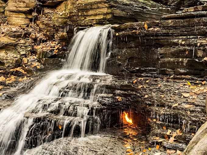 Autumn leaves frame this impossible scene where water cascades over ancient shale while fire burns eternally behind it.