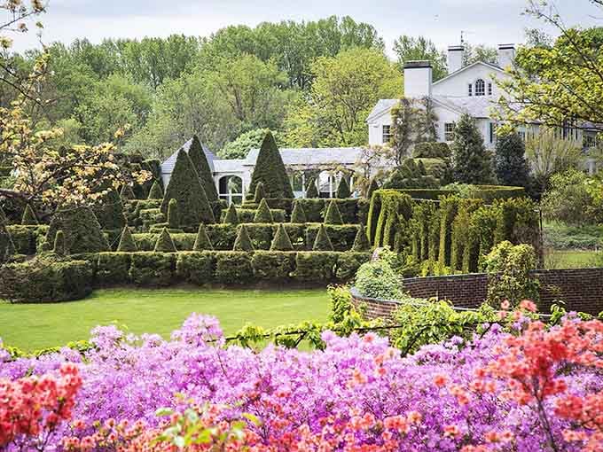The manor house peeks through layers of precisely trimmed topiaries and blooming flowers, blending architectural elegance with natural beauty throughout the property.