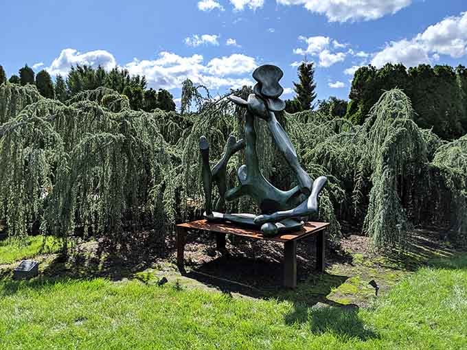 A whimsical bronze figure lounges on a bench beneath weeping willows, living its best sculptural life in perpetual relaxation.
