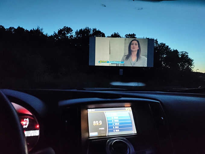 Inside your car, the FM radio display shows 89.9 as the giant screen flickers with pre-show content overhead.
