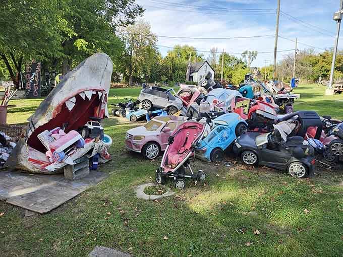 Toy cars and strollers pile together with a shark sculpture, creating unexpected conversations about consumerism and childhood.