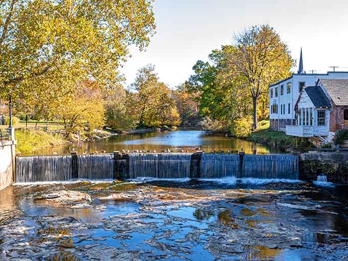Autumn colors reflect in the calm water above the small dam, creating a postcard moment that happens naturally every fall.
