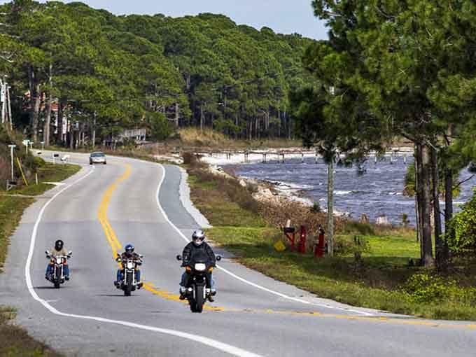 Motorcycles cruise past sparkling Gulf waters where pine forests meet the shore in perfect coastal harmony.