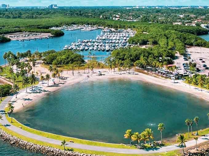 This aerial view reveals a perfect crescent beach and marina where turquoise waters meet tropical palms in stunning harmony.