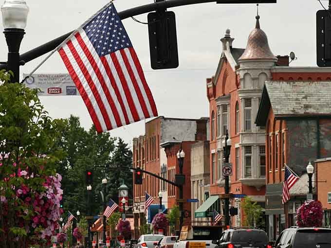 Stars and stripes wave above historic buildings while flowers bloom below, creating pure small-town American perfection.