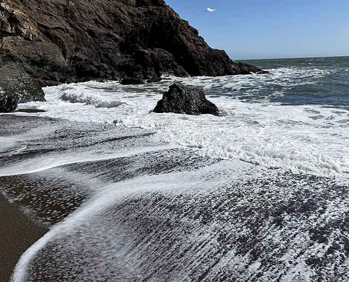 White foam racing across charcoal sand creates nature's own black-and-white photography, no filter needed.