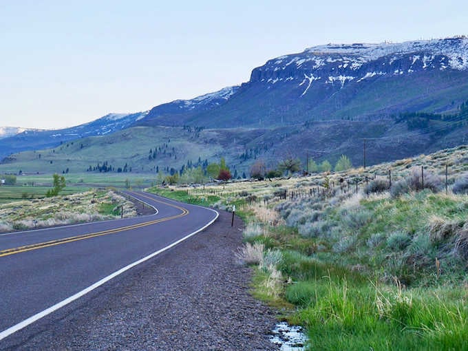 Snow-capped ridges tower above the winding road where sagebrush meets sky in perfect harmony under dramatic clouds.