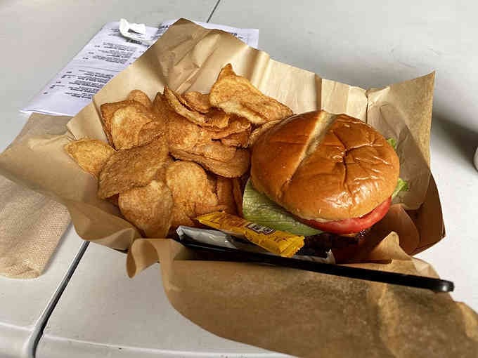 Simple perfection: a golden-toasted bun cradling a juicy burger alongside crispy chips that crunch louder than the gravel outside.