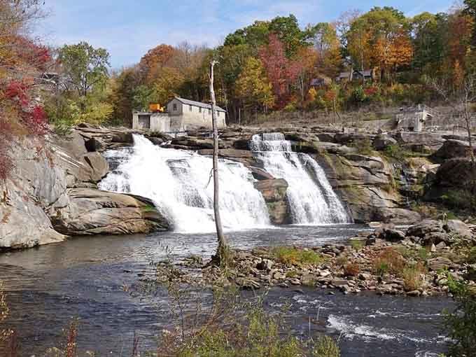 Twin cascades pour over weathered rocks beside historic mill buildings, blending industrial heritage with natural beauty perfectly.
