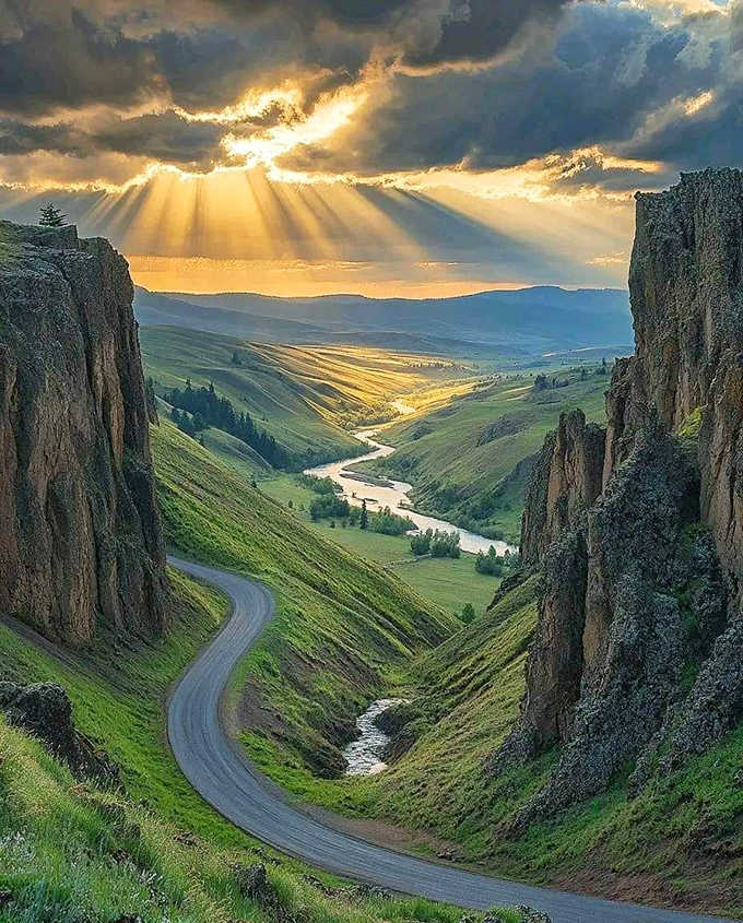Sunbeams pierce storm clouds like spotlights on a stage, illuminating green valleys carved by ancient rivers and time.